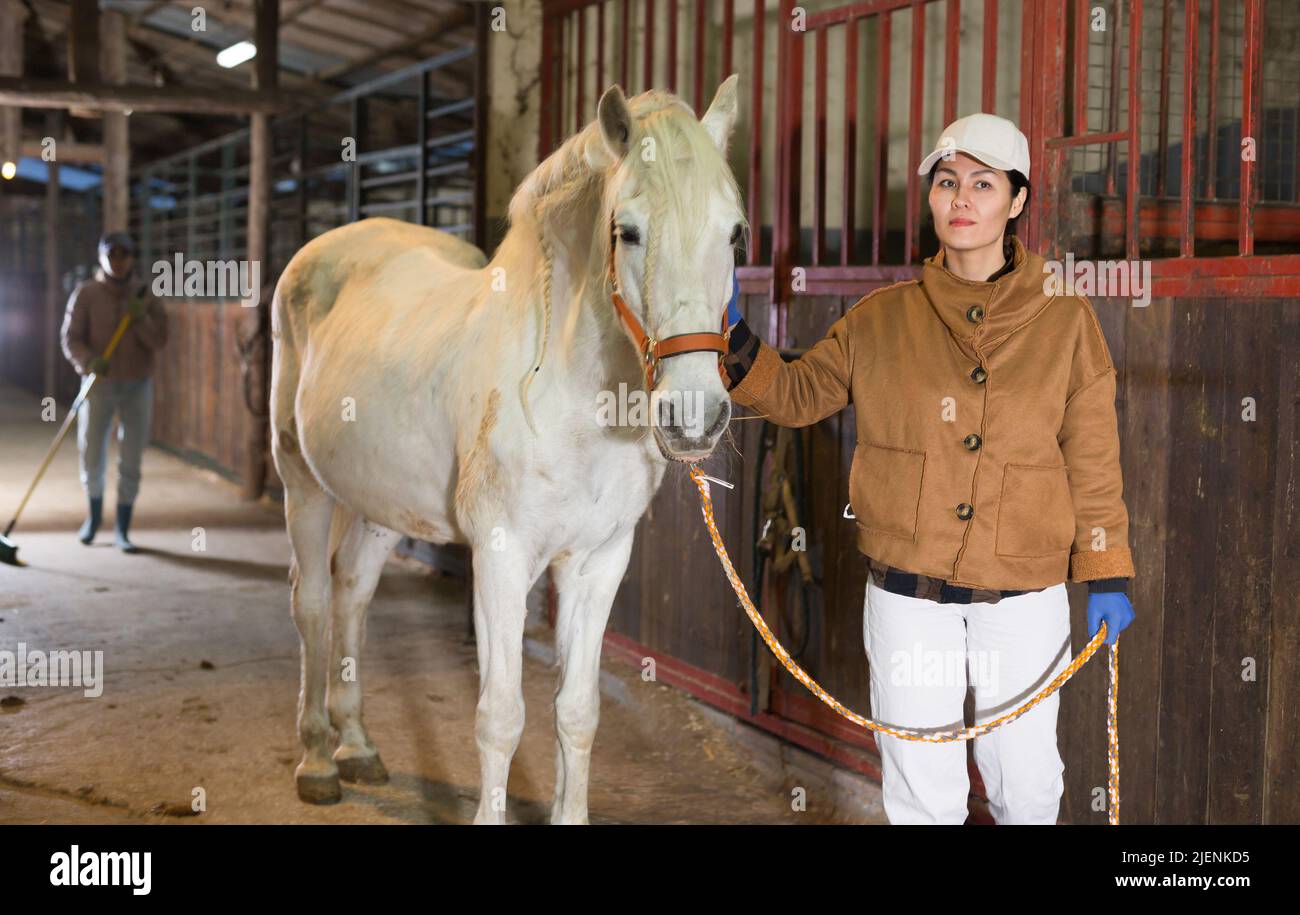 Asian female stable worker leading horse by bridle in barn Stock Photo ...