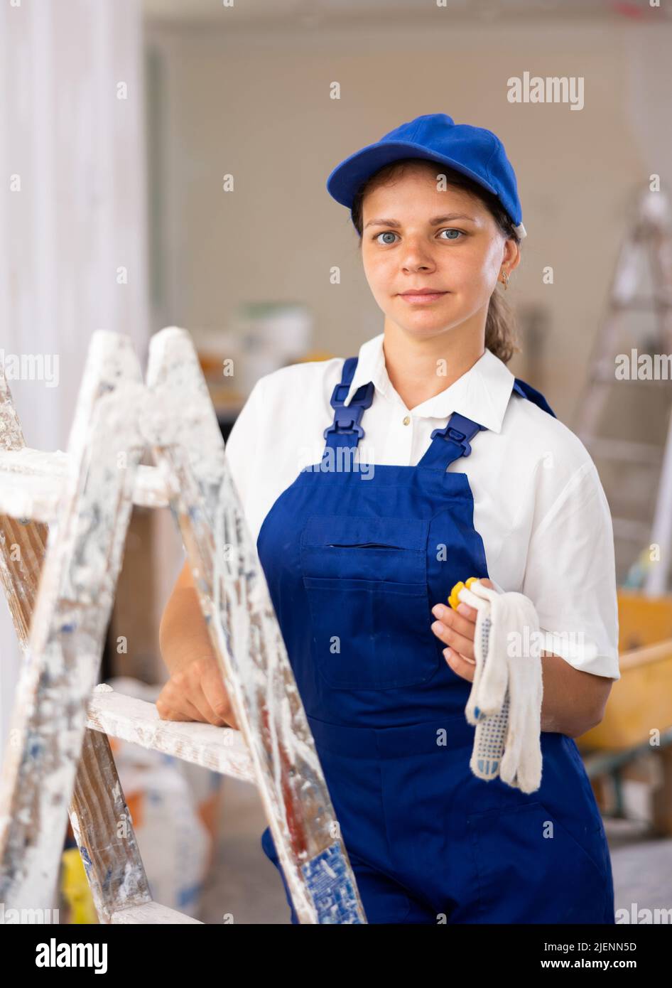Portrait of positive builder woman in blue overalls next to stepladder ...