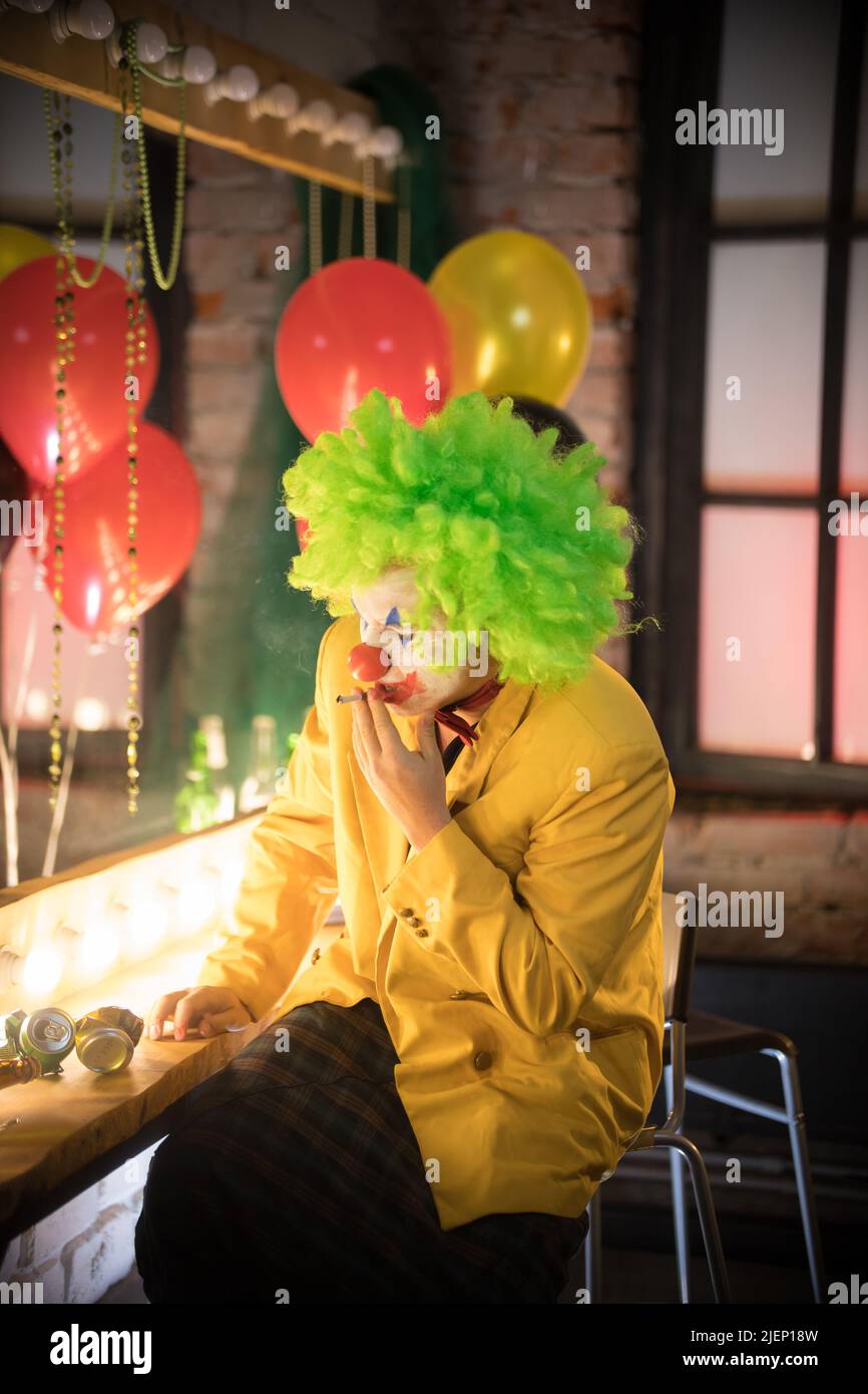 A sad tired man clown in yellow jacket and green wig sitting in the ...