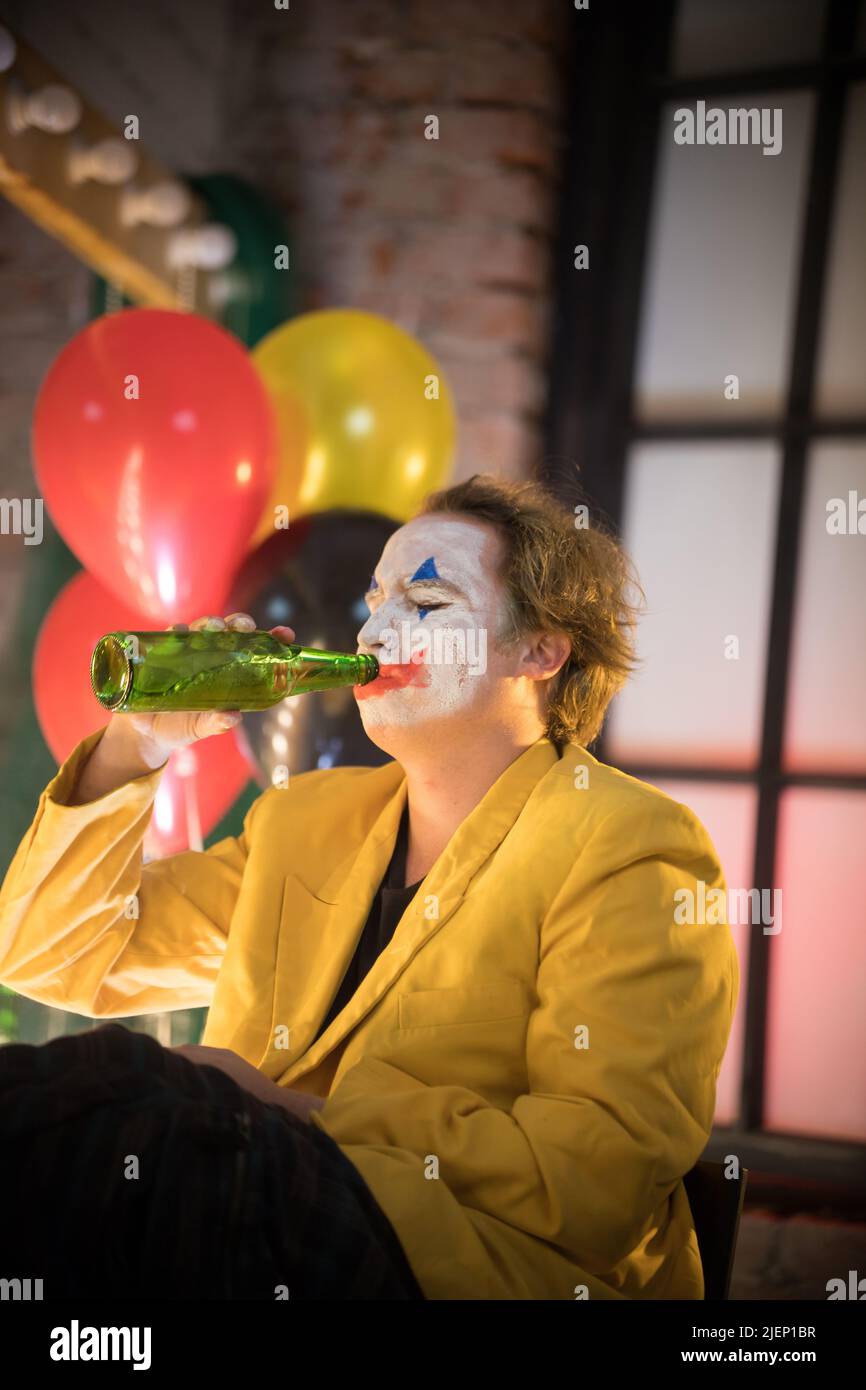Clown concept - a sad tired man clown in yellow jacket drinking alcohol in the dressing room ...