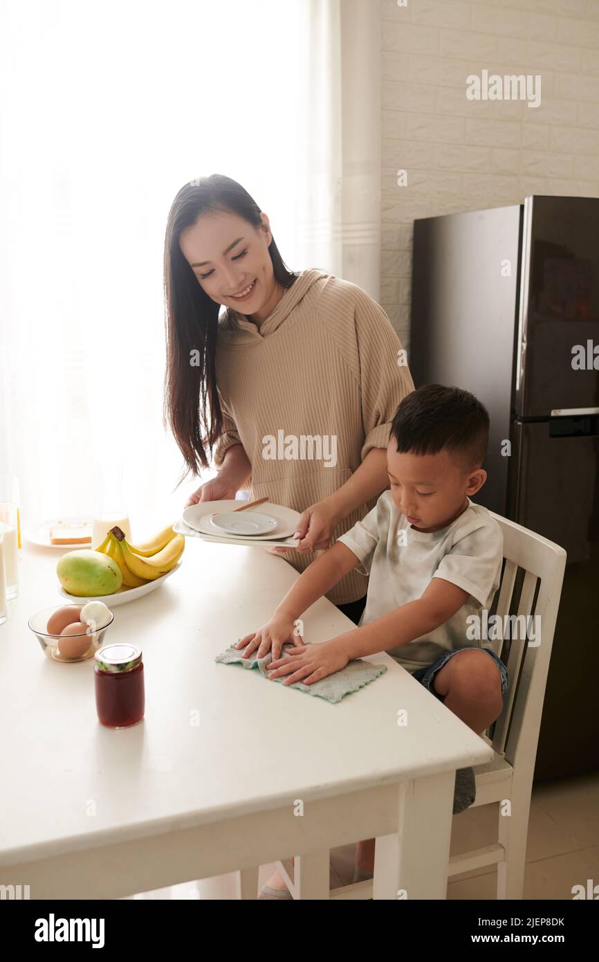 Smiling little boy wiping kitchen table with soft cloth after breakfast ...