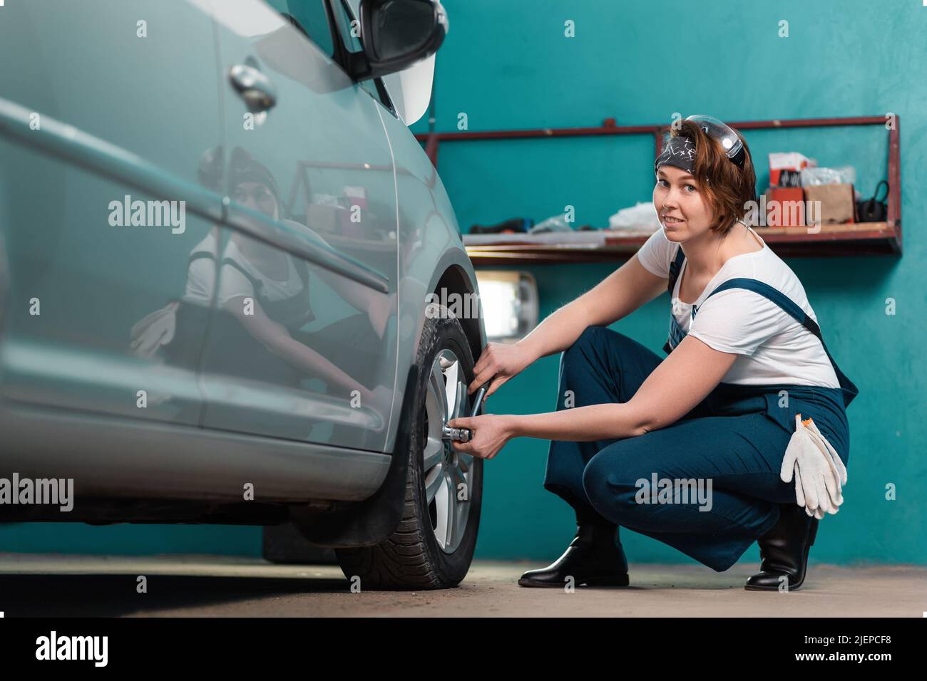 Garage work. Young smiling woman mechanic in blue coveralls squatting ...