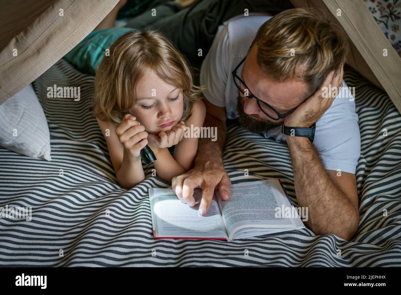 Portrait of a 6 year old boy and his father reading a book in teepee ...