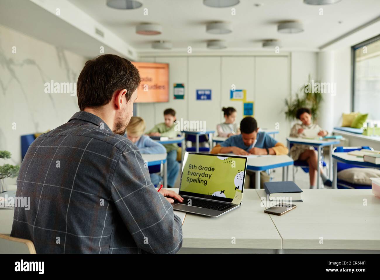 Back view at male teacher using laptop at desk in classroom with group ...