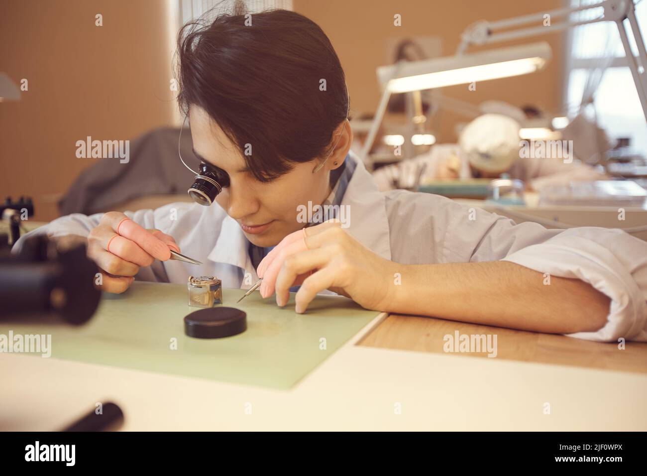 Concentrated young female watchmaker in lab coat sitting at table and ...