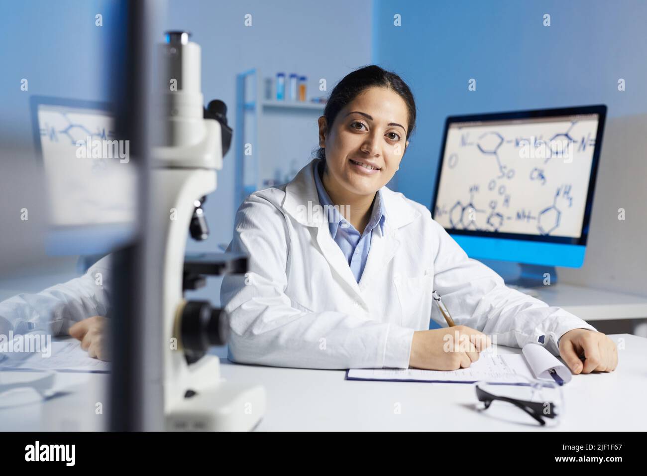 Portrait of smiling young Indian woman in lab coat sitting at desk and ...