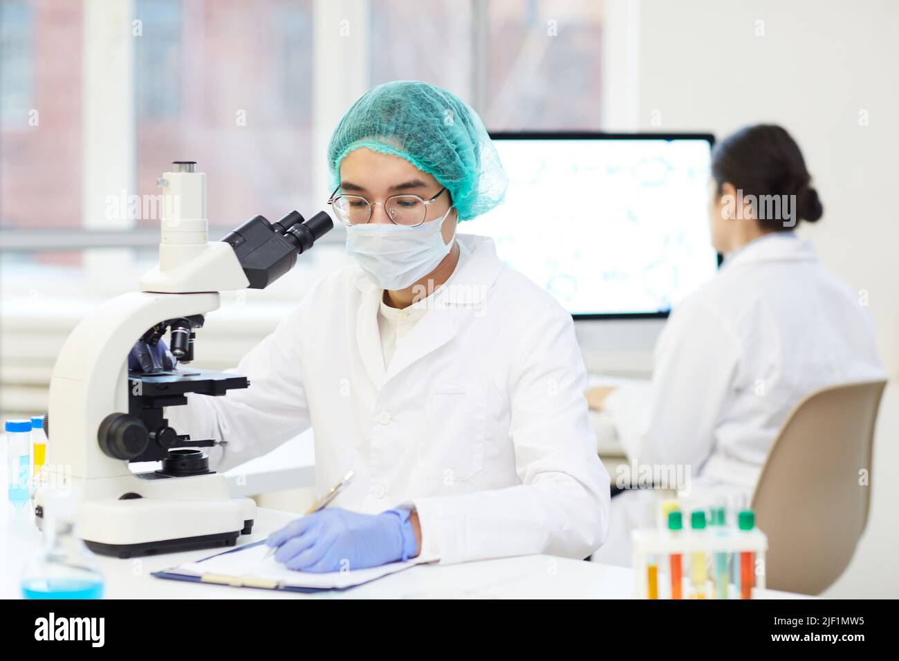 Serious young Asian laboratory researcher in sterile workwear sitting ...