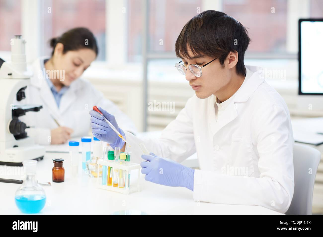 Serious young Asian lab technician in protective gloves and white coat ...