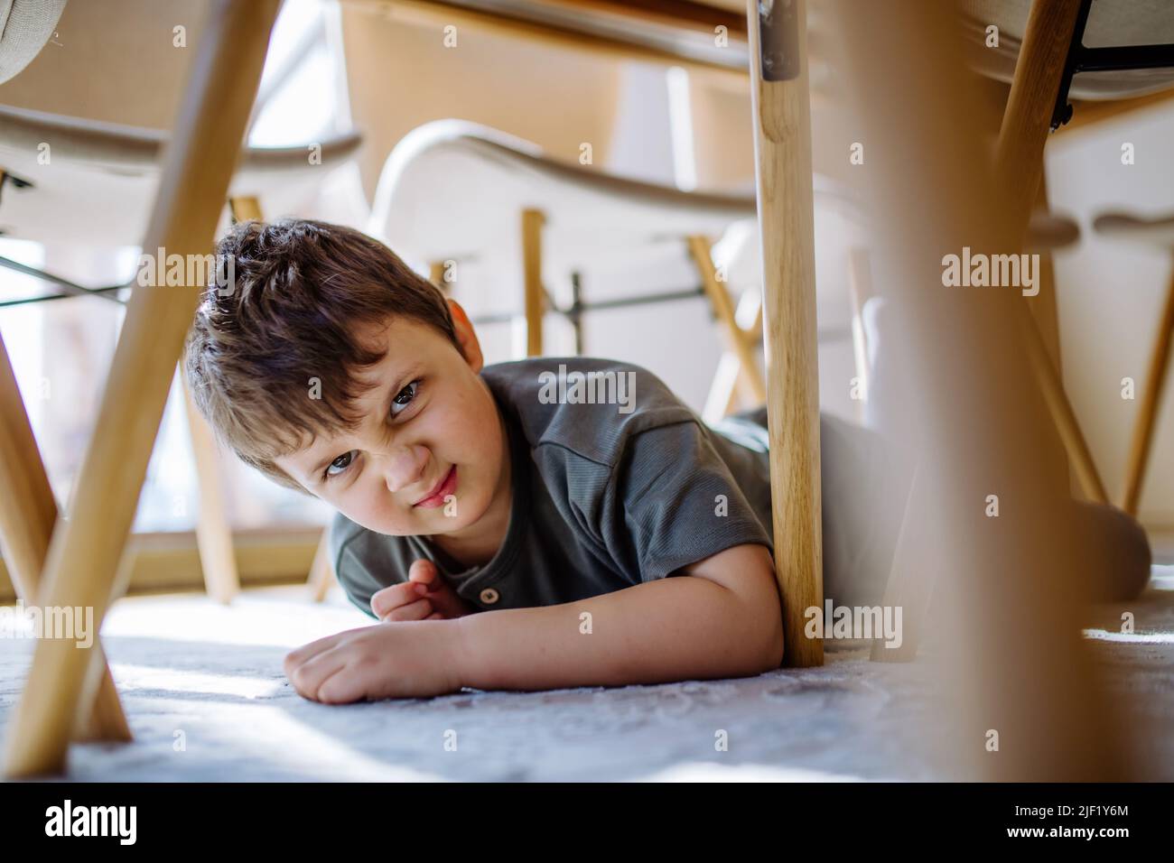 A lttle boy hiding under the chair at home and making angry grimace ...
