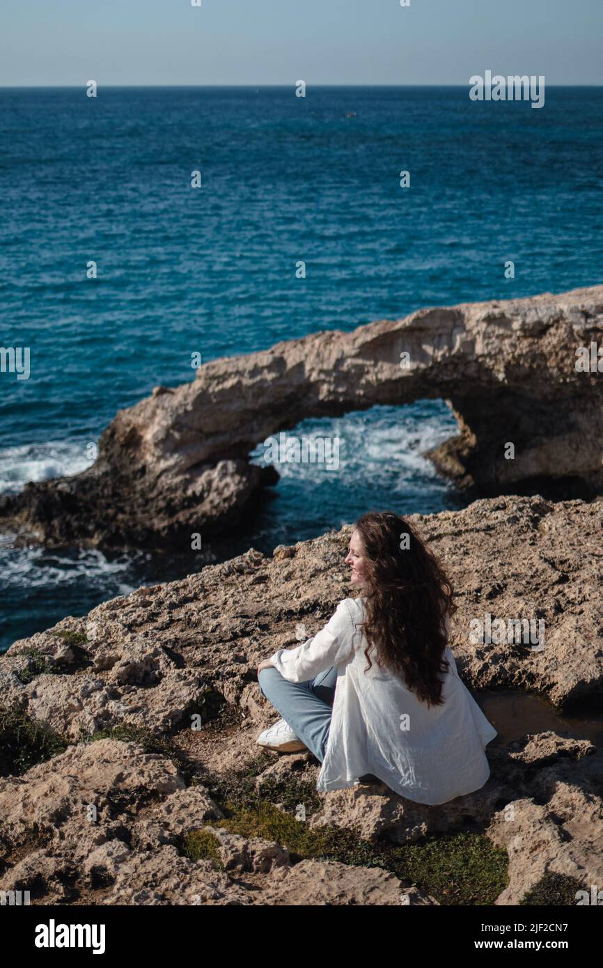 Relaxed woman from behind sit at the edge and looking at sea cave arch ...