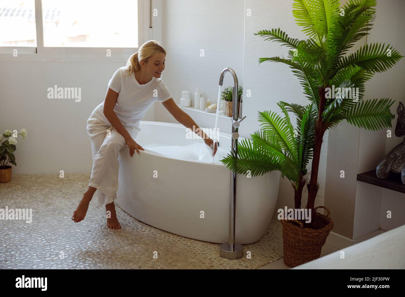Blonde young beautiful female preparing warm bath with bubbles and ...