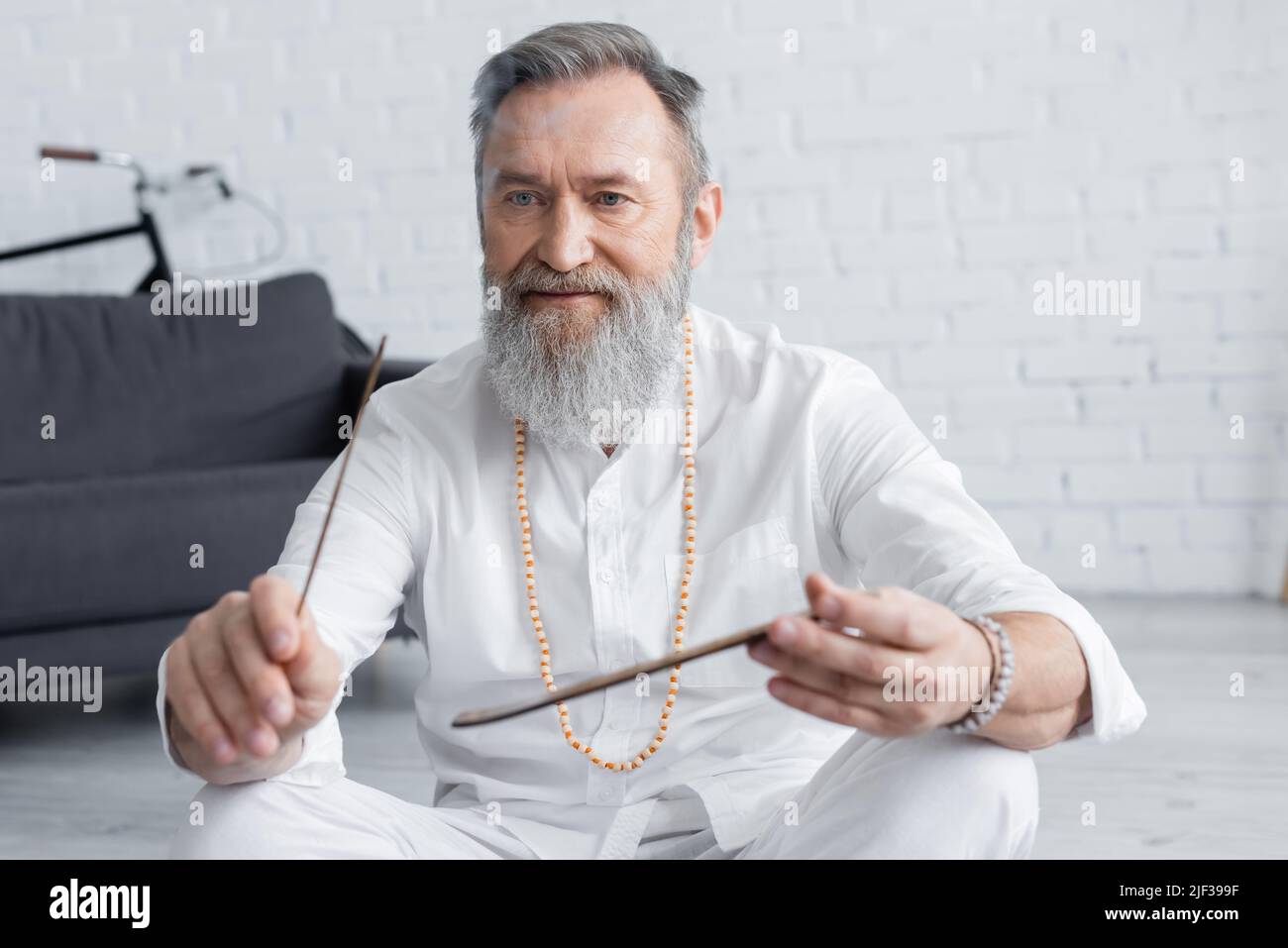 senior guru man in white shirt and beads holding aroma stick at home ...