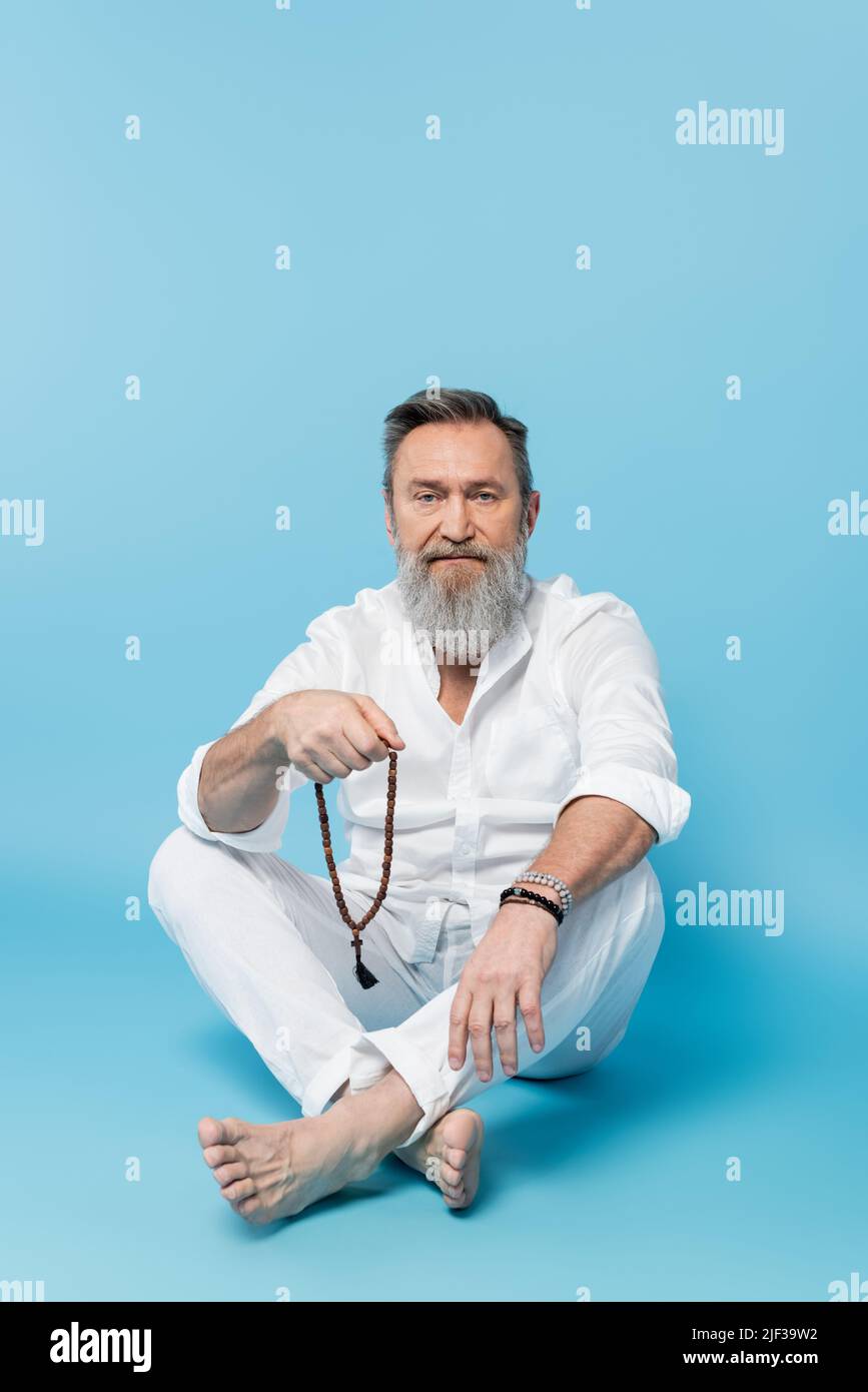 bearded barefoot man sitting with crossed legs and holding mala beads ...
