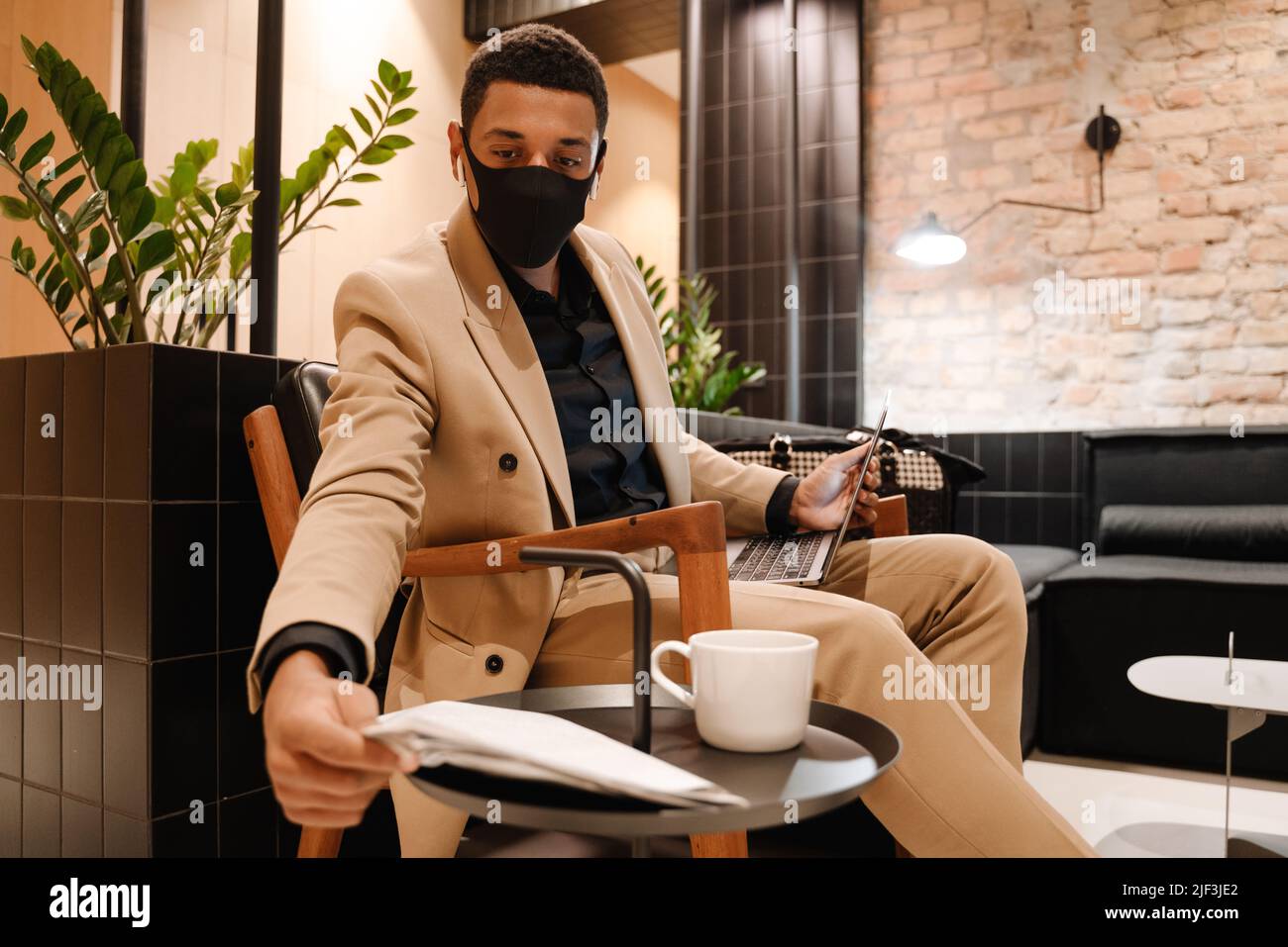 Black man in face mask reading newspaper while working with laptop at ...