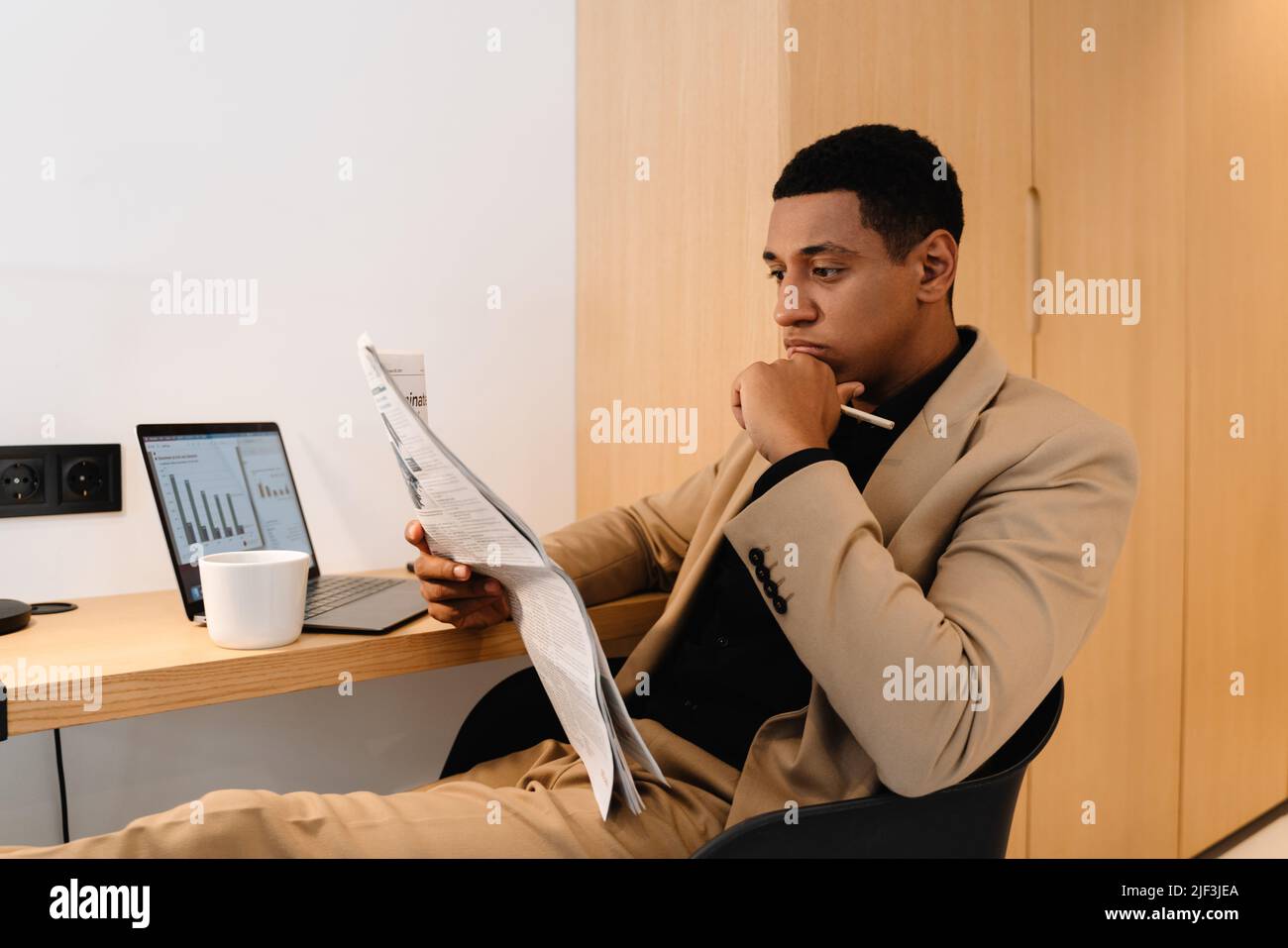 Black man reading newspaper while working with laptop at desk in hotel ...