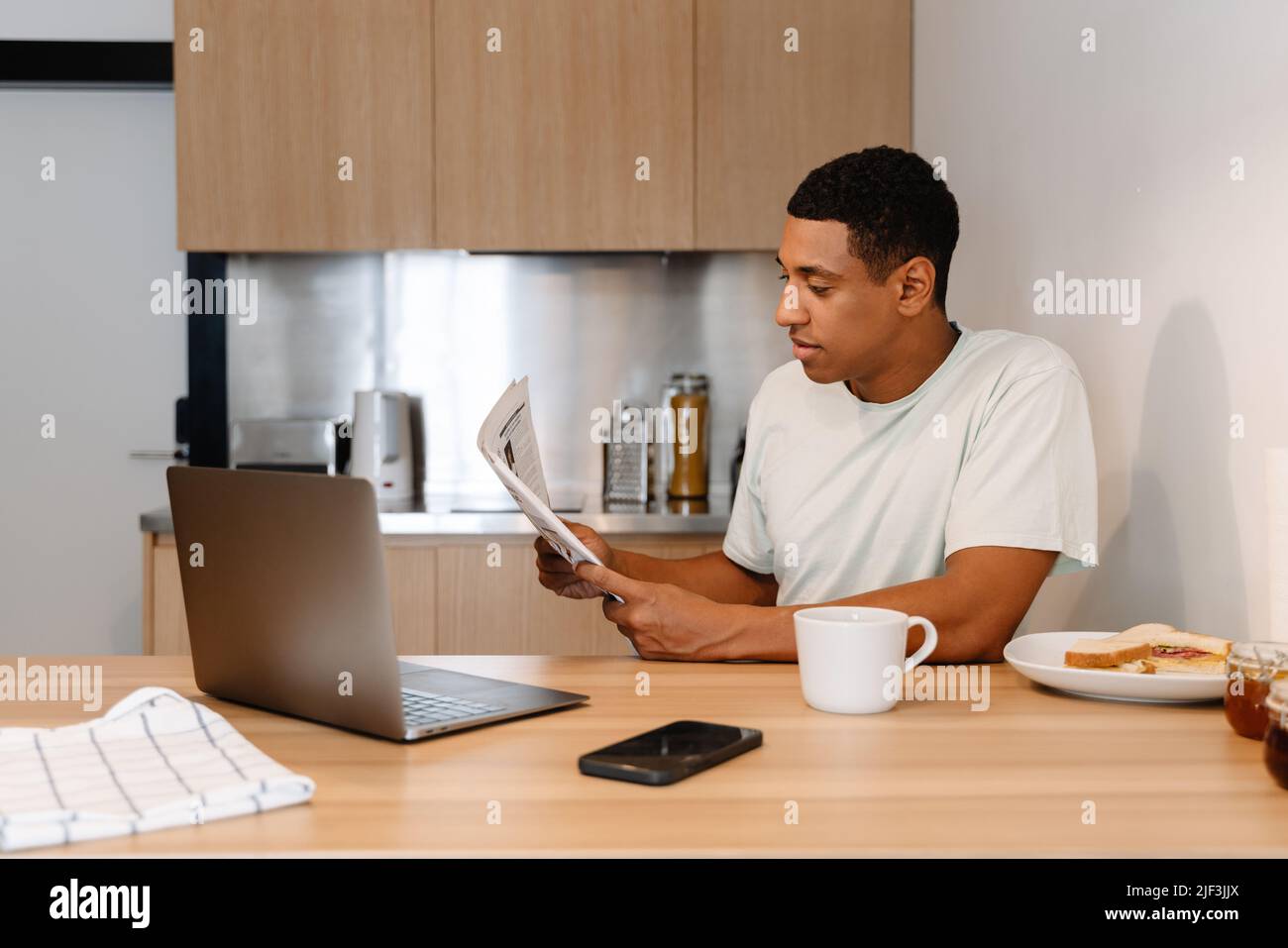 Black man reading newspaper and using laptop while having breakfast in ...