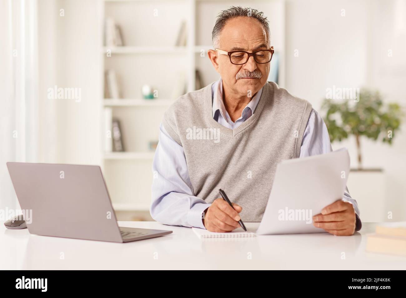 Mature man sitting in front of a laptop computer looking at a paper ...