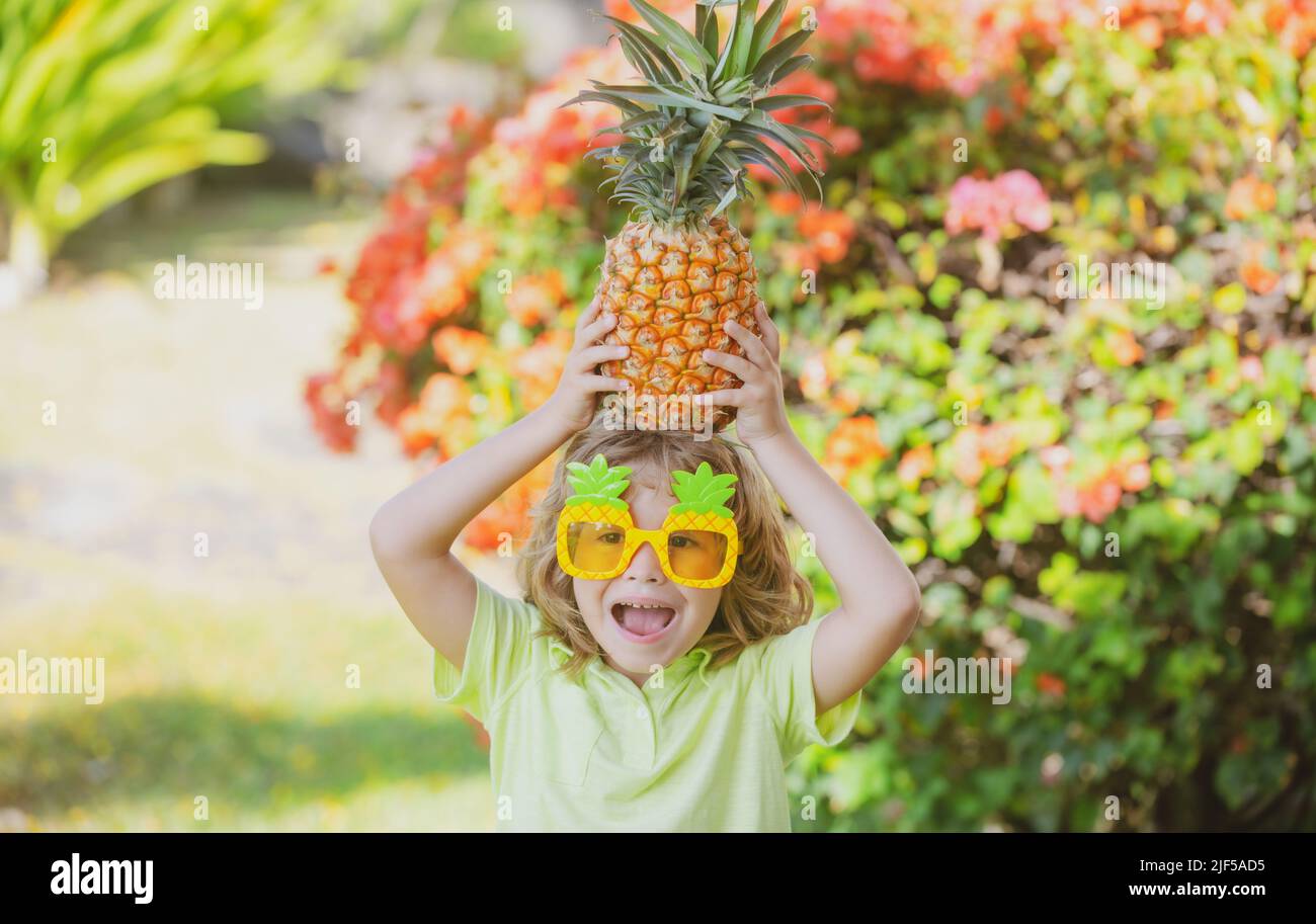 Funny child holds a pineapple on her head on a summer nature background. Pineapple kid boy and ...