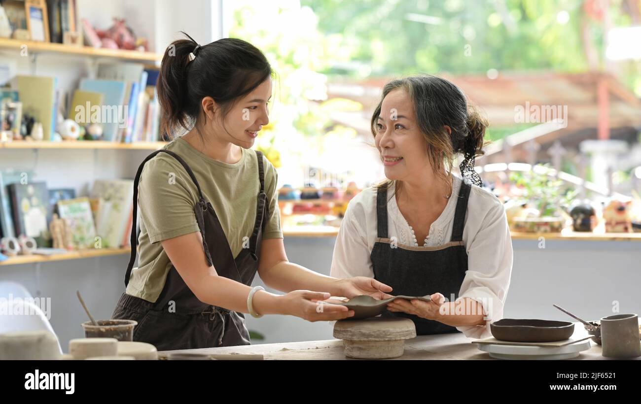 Young asian woman teaching mature woman making ceramics in pottery ...