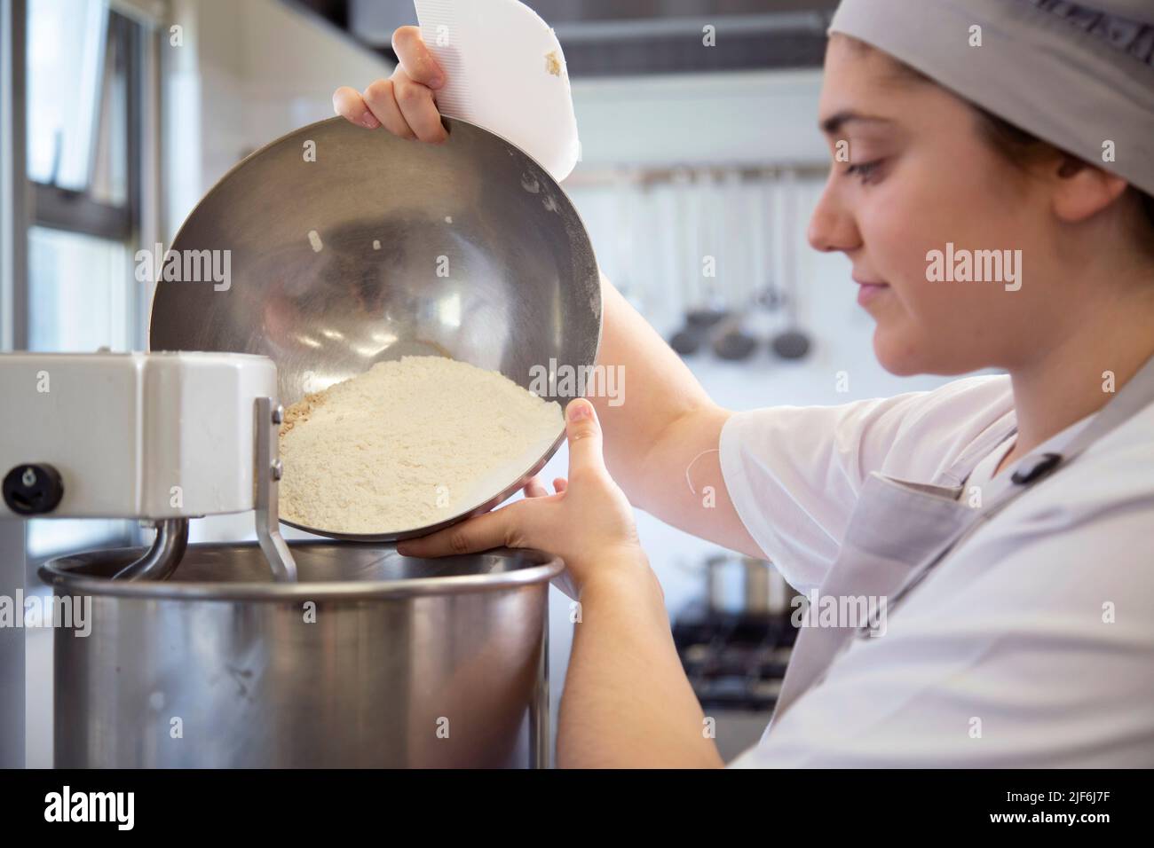 Side view of crop female baker in uniform pouring flour into bowl of ...