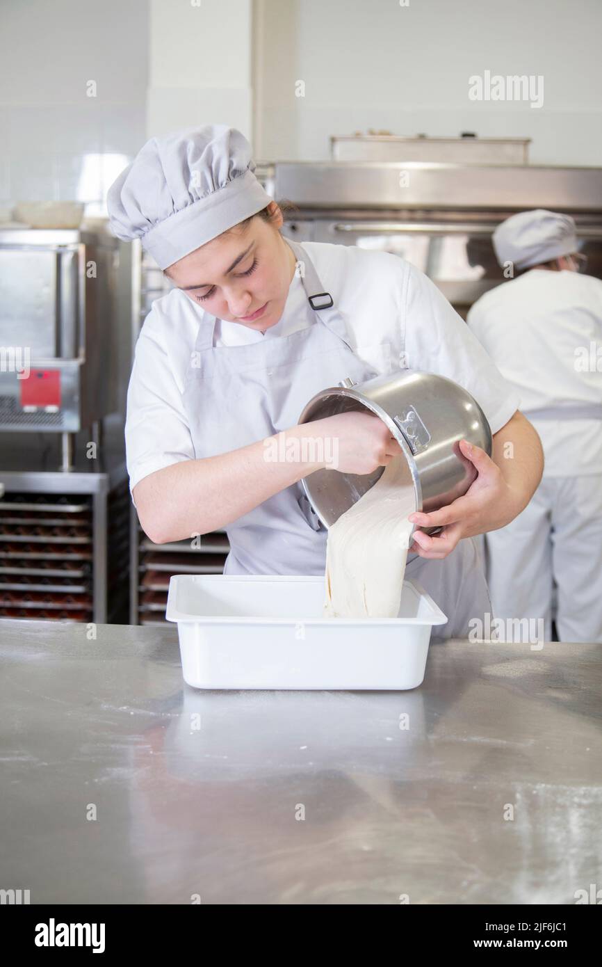 Side view of female baker in uniform putting raw dough from bowl into ...