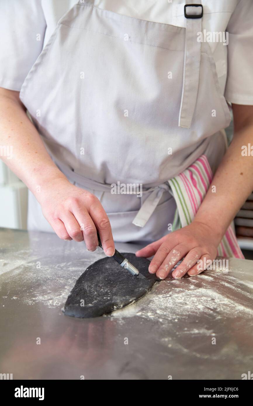 Crop anonymous female baker cutting raw black dough with knife on messy ...