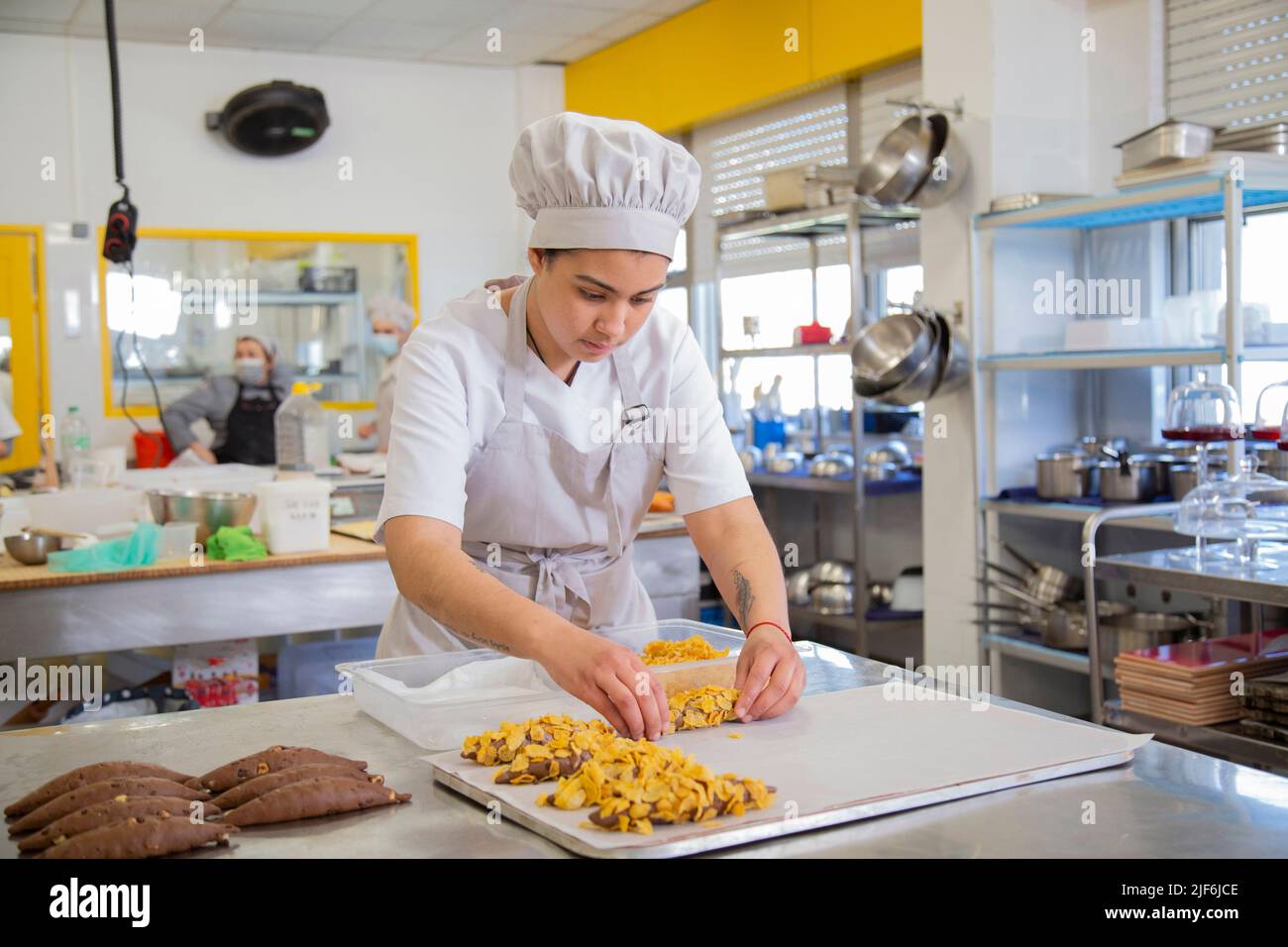 Focused female cook in uniform covering cookie dough with cornflakes at ...