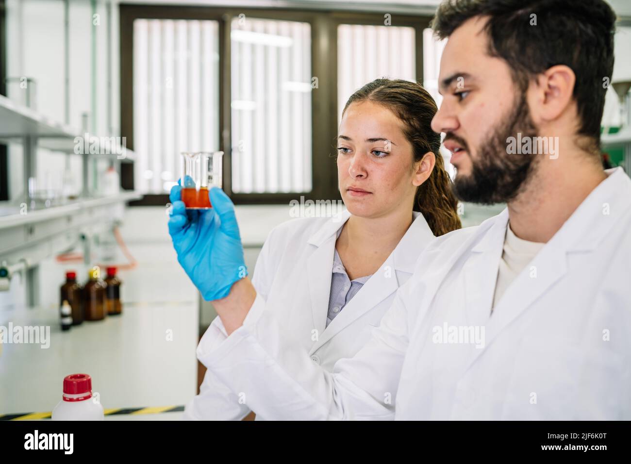 Attentive scientists in medical gowns examining beaker with chemical ...