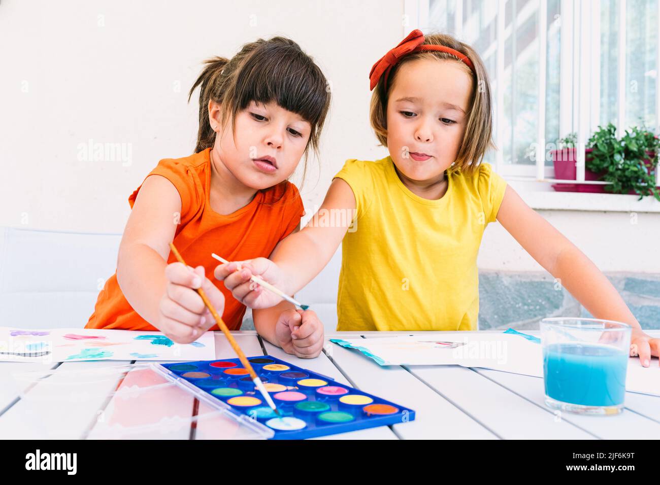Cute girls painting with paintbrushes on papers at table with palette ...