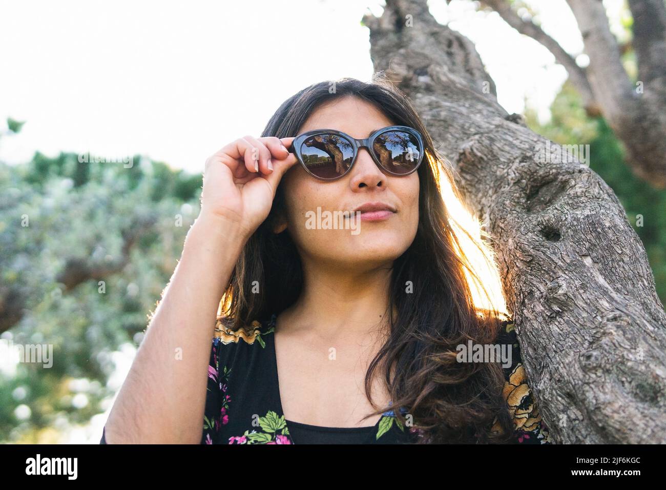 Female in sunglasses looking away in back lit leaning on tree trunk on ...