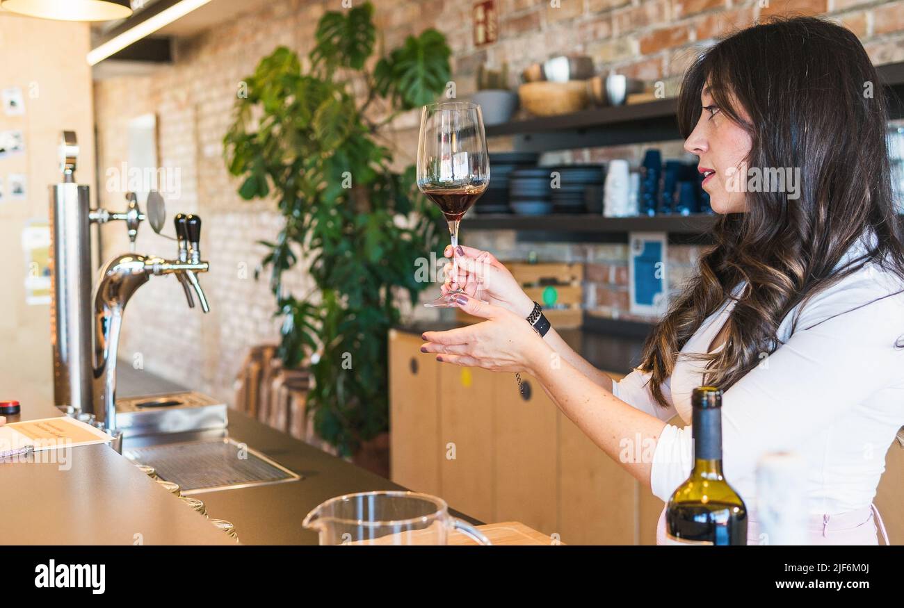 Side view of young brunette swirling red wine in glass goblet to ...