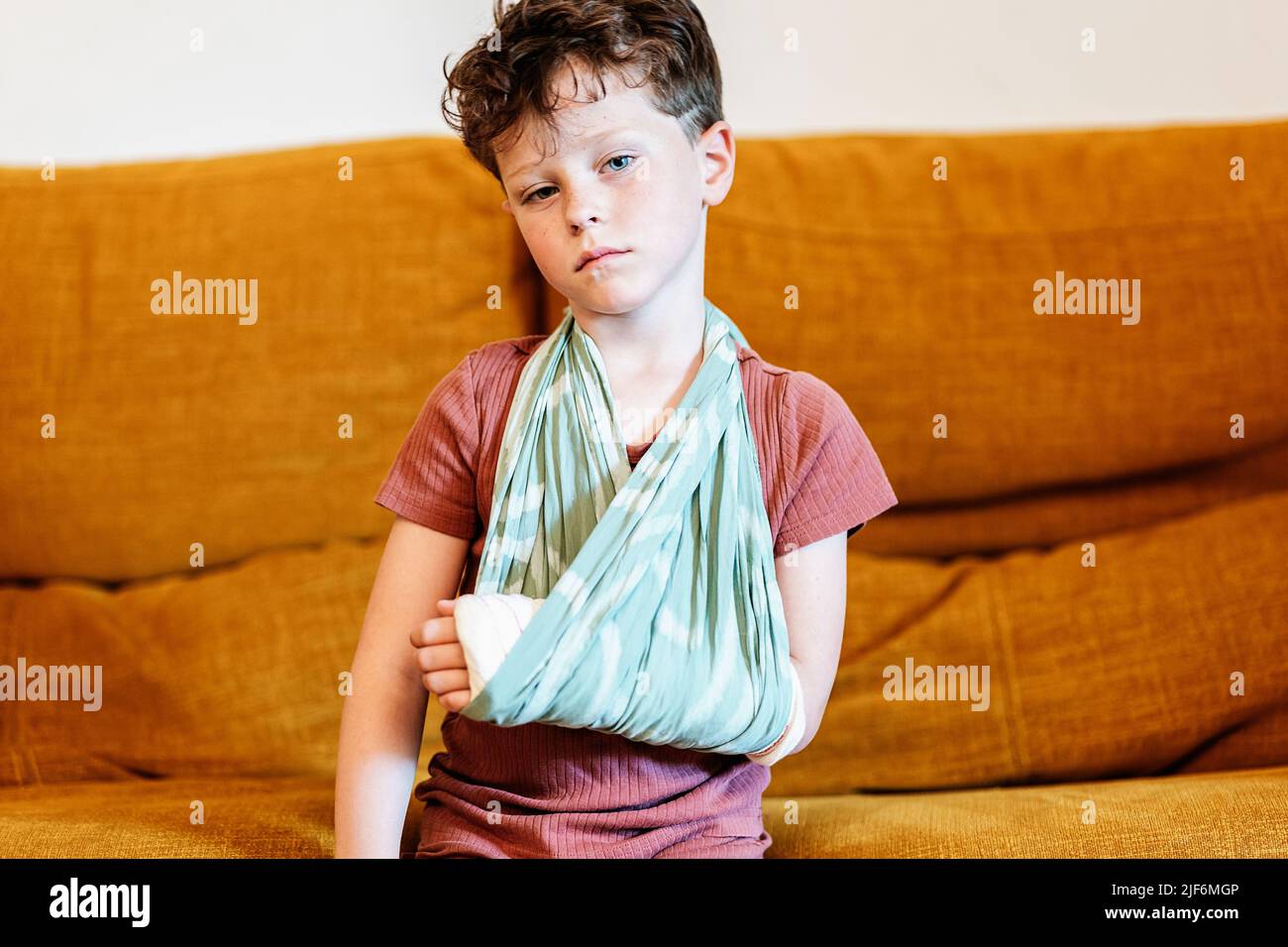 Little boy in t shirt caressing broken arm in sling looking at camera ...