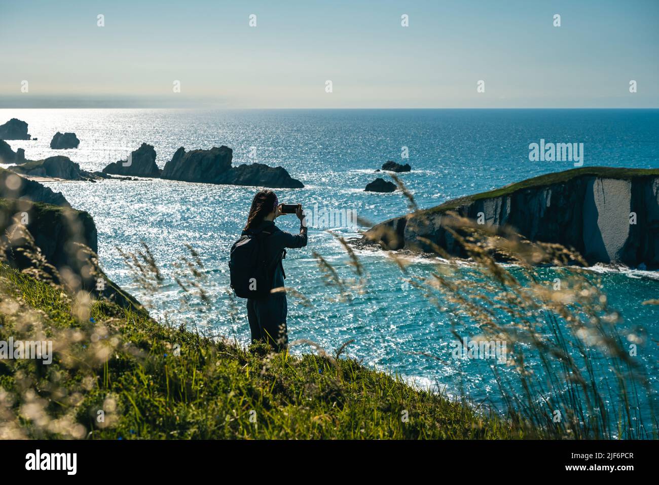 Rear view of female hiker photographing cliff using smartphone near sea ...