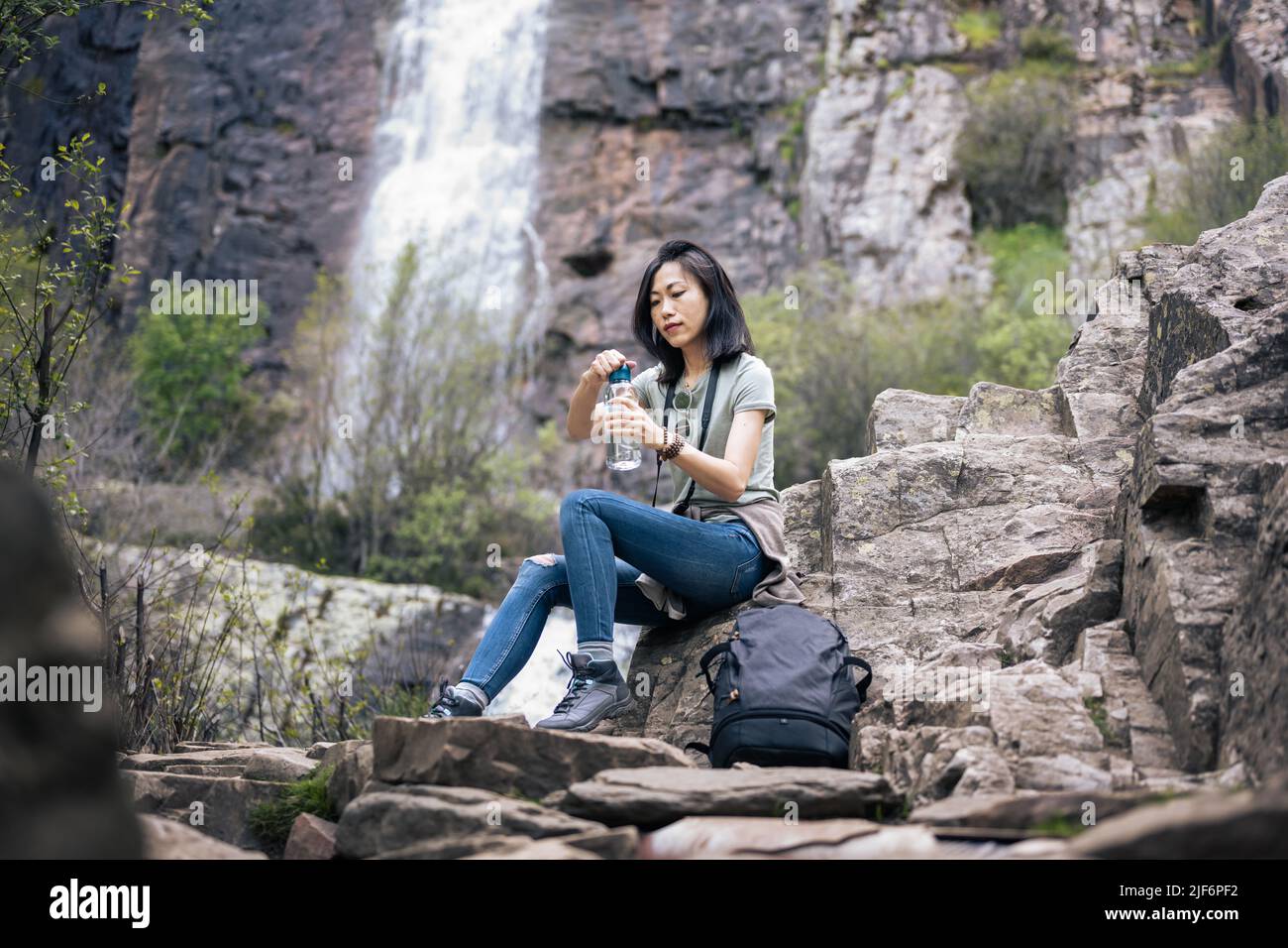 Full body of Asian female hiker opening bottle of water sitting near ...