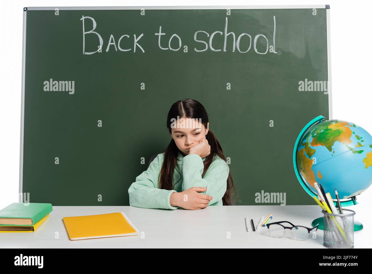Sad pupil sitting near books on table and chalkboard with back to ...
