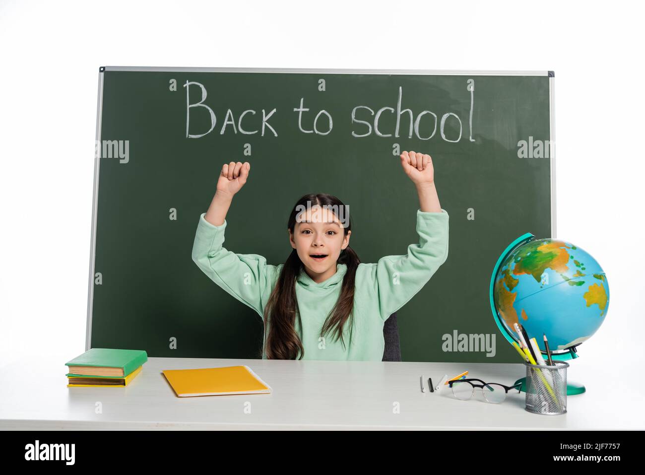 Excited schoolgirl sitting near books and chalkboard with back to ...