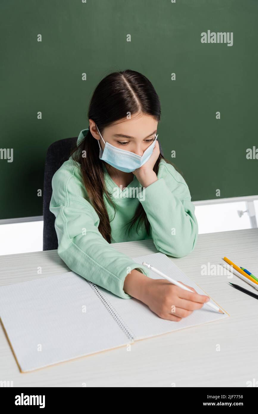 Schoolkid in medical mask writing on notebook near chalkboard isolated ...