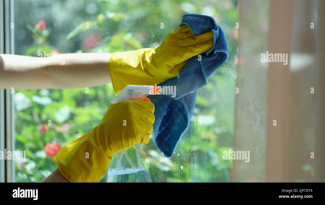 Woman hand in yellow gloves washes window with blue rag and detergent ...