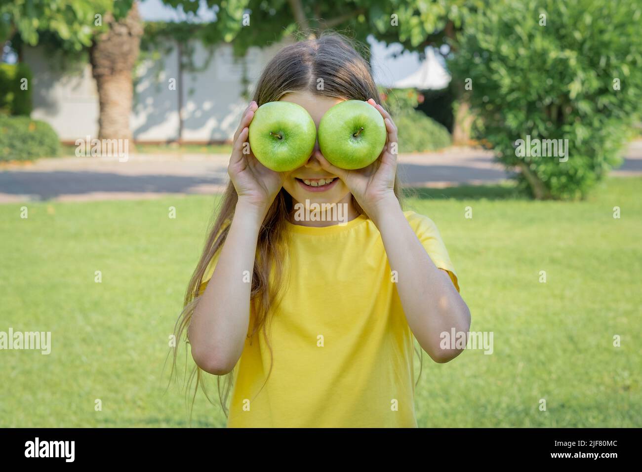 A teenage girl holds two green apples in front of her face in the park ...