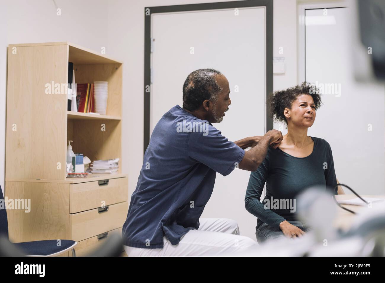 Male healthcare worker examining shoulder female patient grimacing with ...