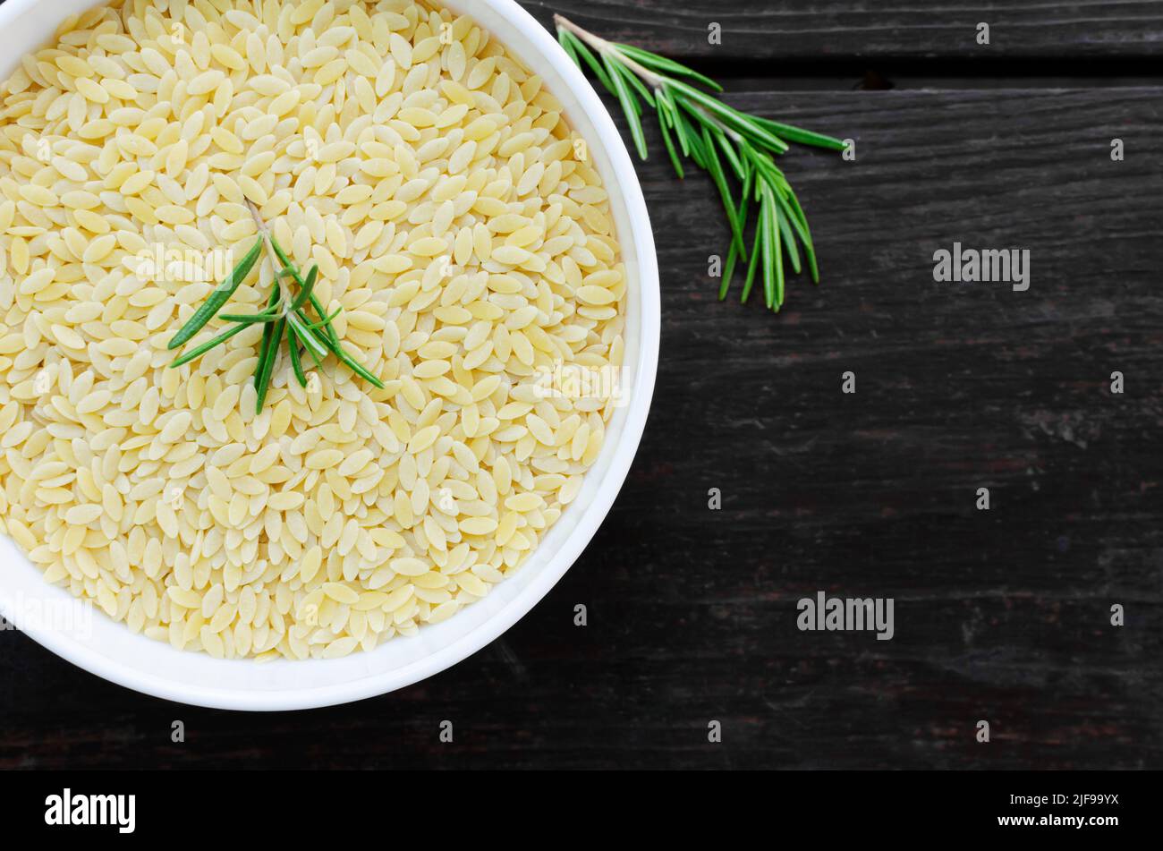 Raw risoni pasta in a white bowl with a sprig of rosemary on a wooden ...