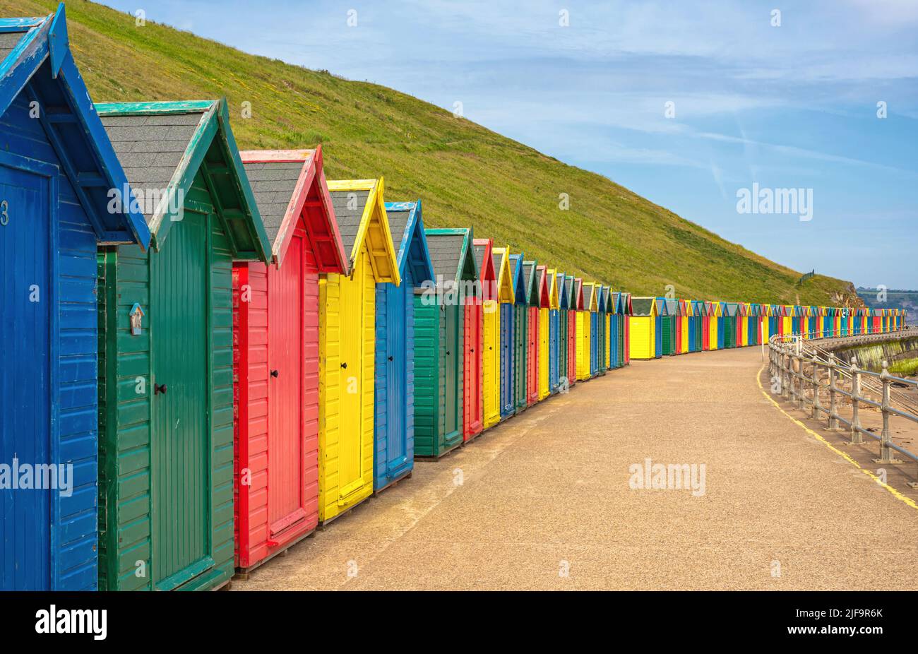 A line of colourful beach huts at the base of a grass covered hill ...