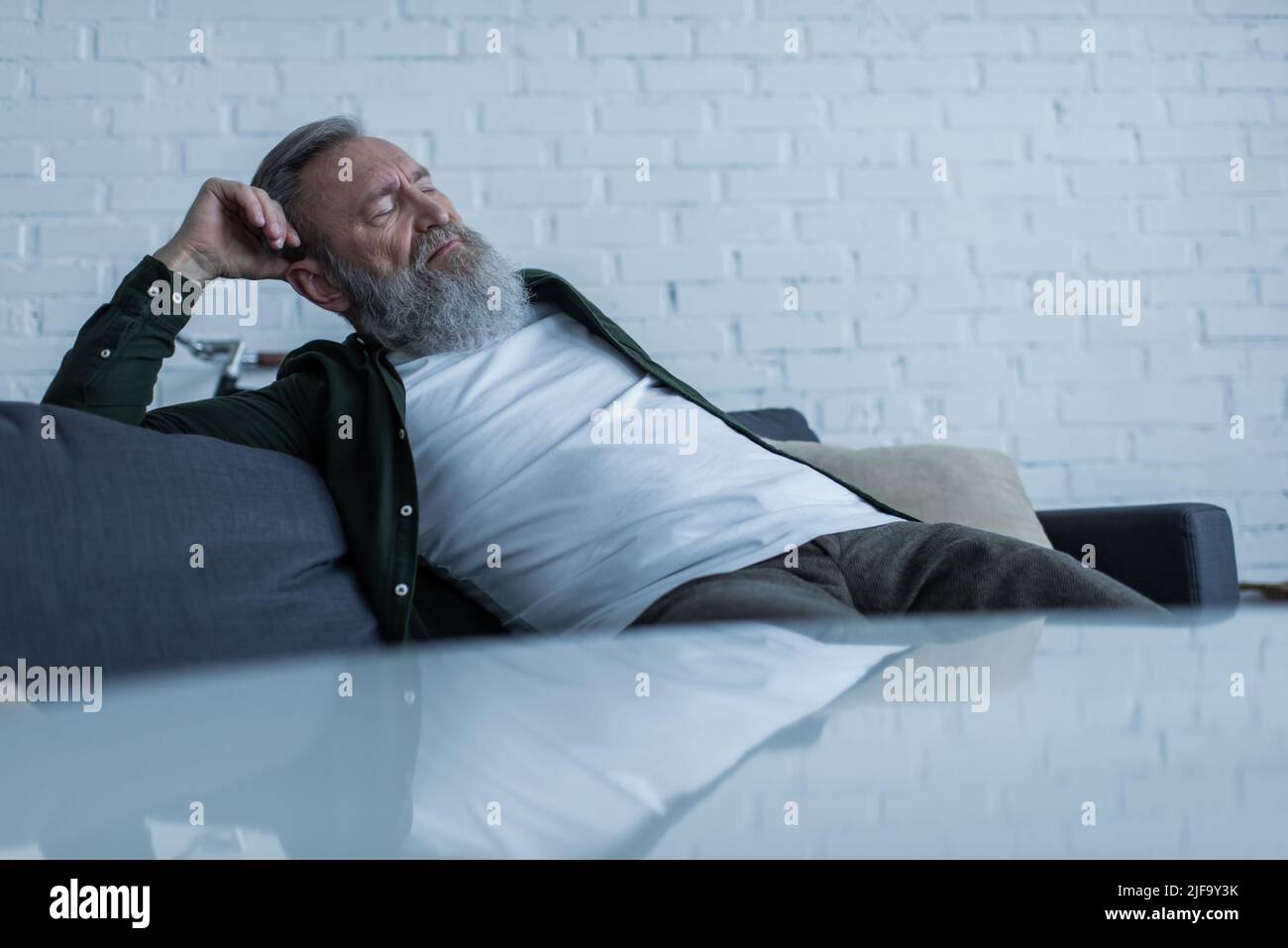 upset senior man with beard suffering crisis while sitting on couch in ...