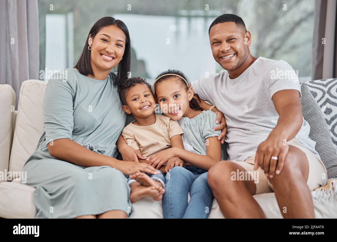 Happy and affectionate young mixed race family of four sitting on a sofa in the living room at ...