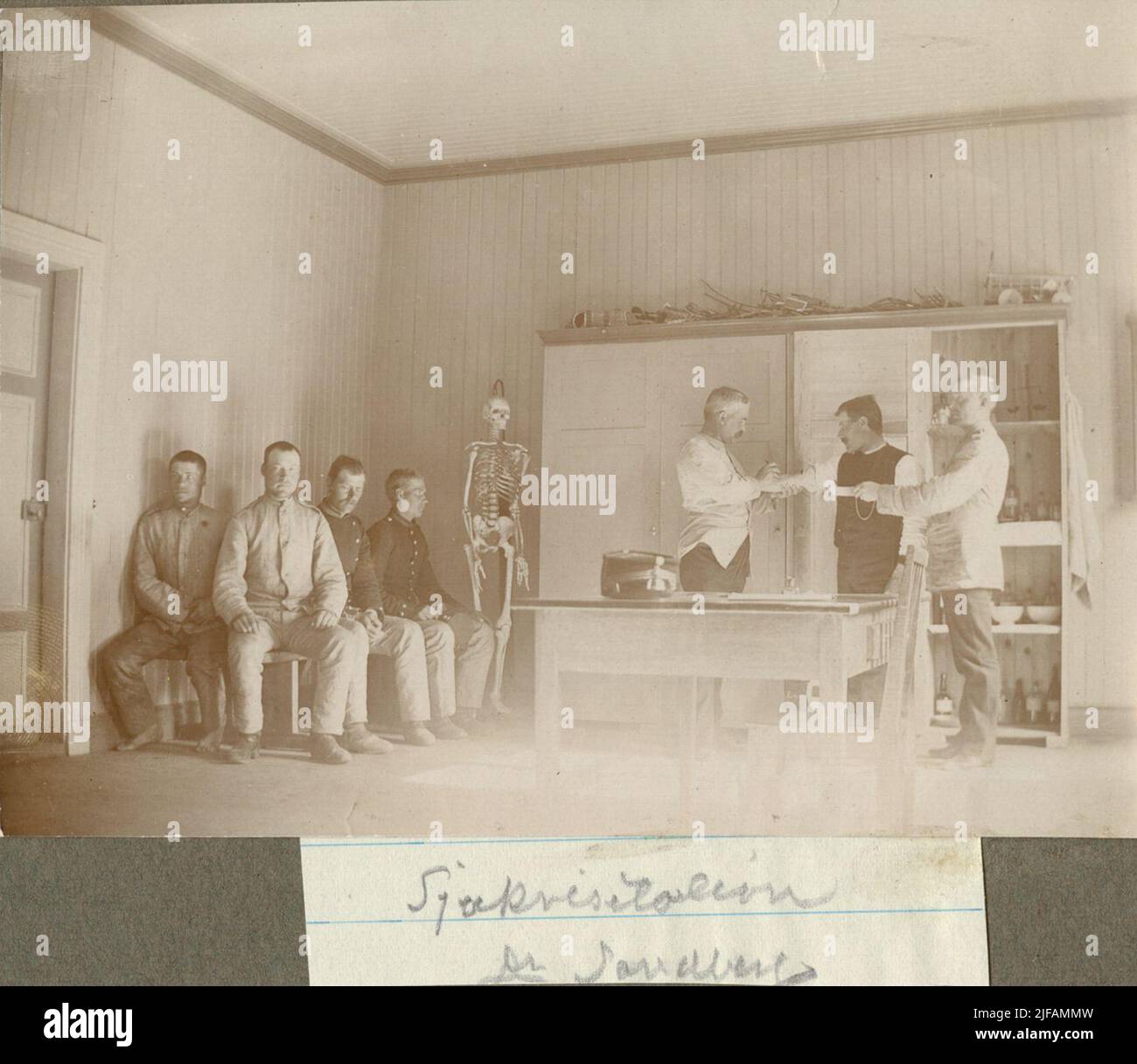 Doctor Sandberg with assistant examines a patient. Four soldiers sit on ...