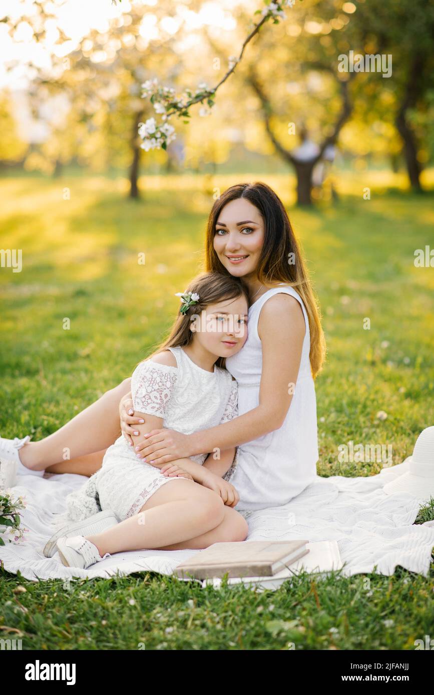 Mom hugs her daughter, sitting on a blanket in a spring blooming garden. Maternal love for a ...