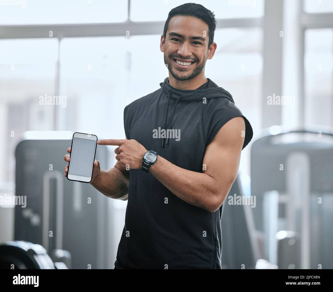 Portrait of smiling asian trainer alone in gym showing cellphone screen ...