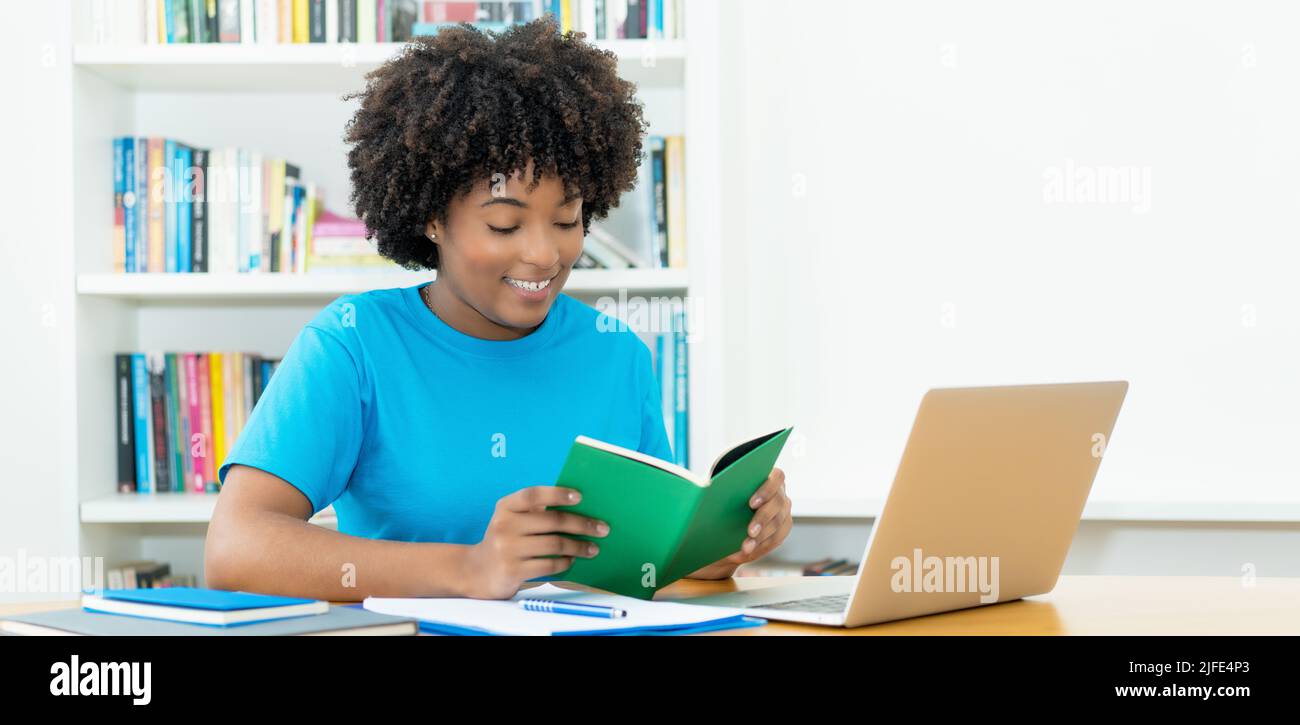 Laughing african american female student reading reference book at desk at library Stock Photo ...