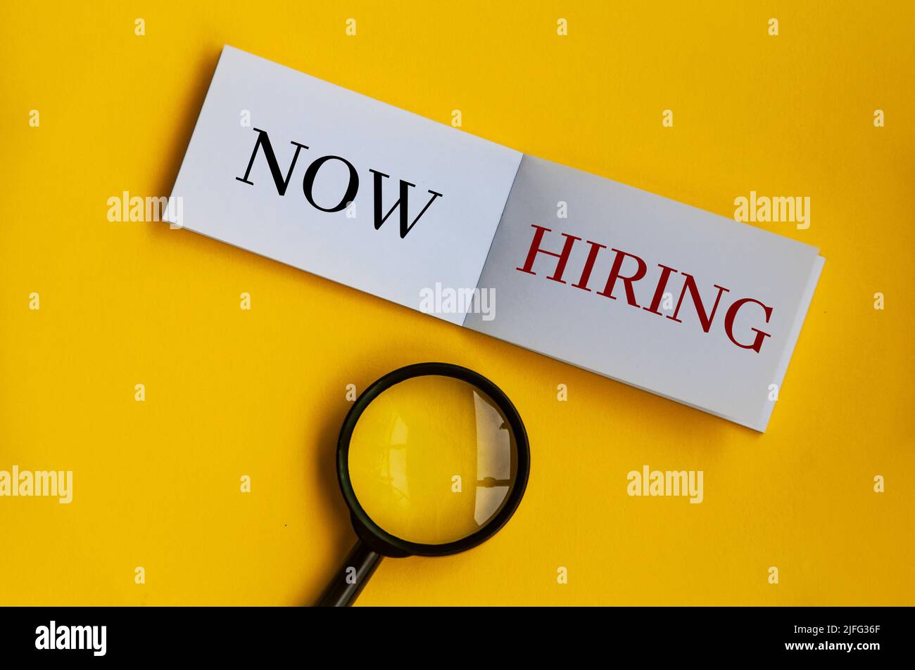 Now hiring text on notepad with magnifying glass on yellow background ...