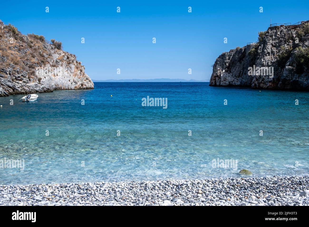 Greece. Pebble beach, moored boat in transparent calm sea, blue sky ...